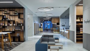 Um saguão moderno de hotel com balcão de recepção, área de estar e cozinha visível ao fundo.