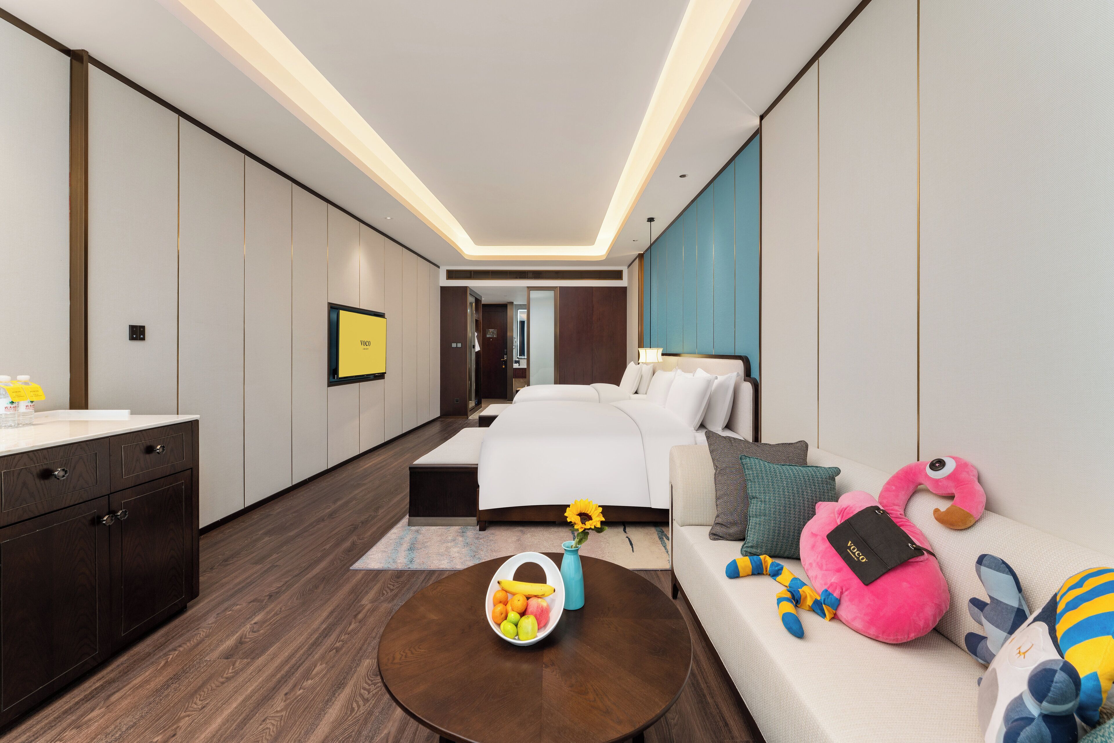 Quarto de hotel moderno com uma cama grande, um sofá com brinquedos macios, uma mesa de centro com frutas e um banheiro visível através de uma porta aberta.
