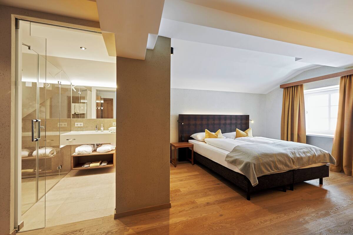 Quarto de hotel moderno com chuveiro envidraçado, cama grande com cabeceira, mesa de cabeceira e vista para o exterior.