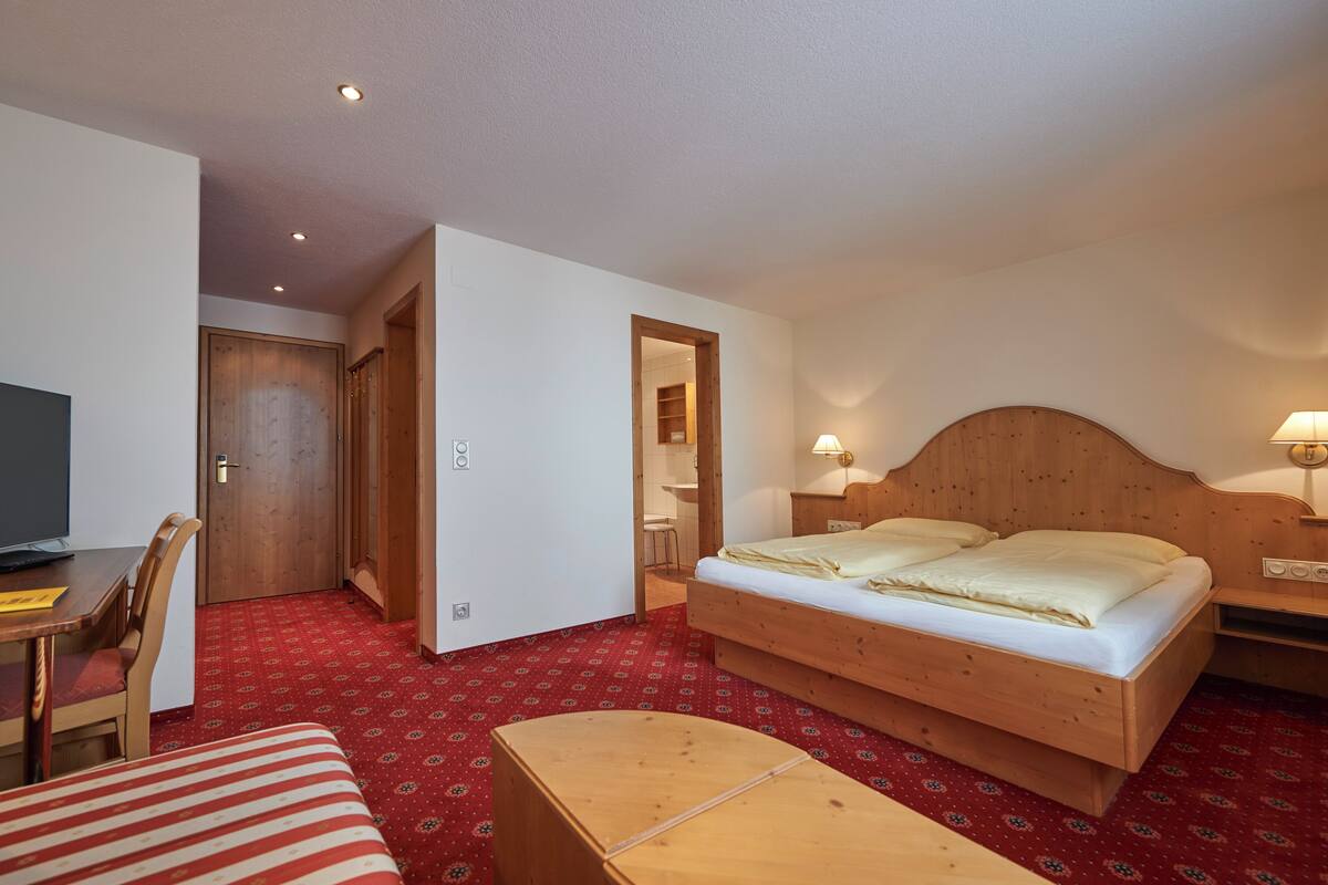 Quarto de hotel com uma cama de madeira, uma escrivaninha com cadeira, uma mesinha e um tapete vermelho estampado.