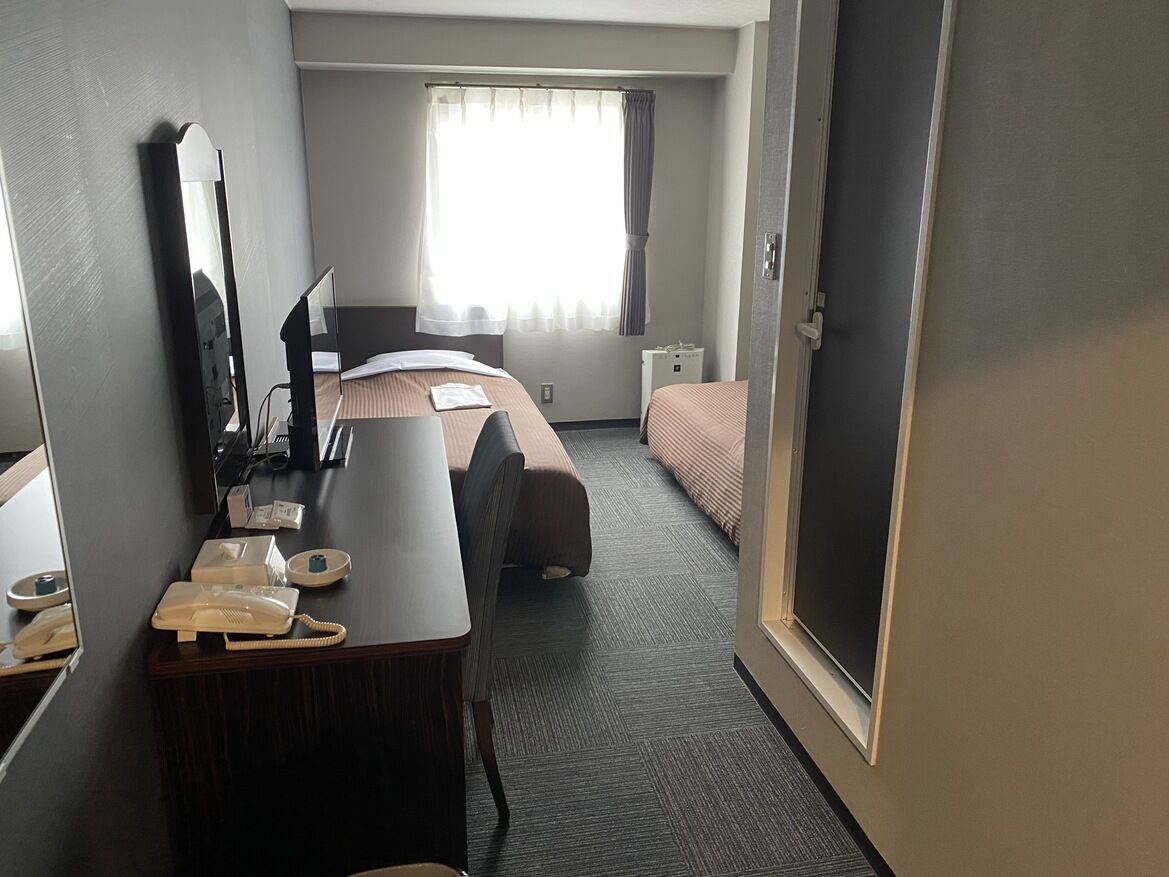 Quarto de hotel com escrivaninha, cama, espelho, telefone e janela com cortinas.