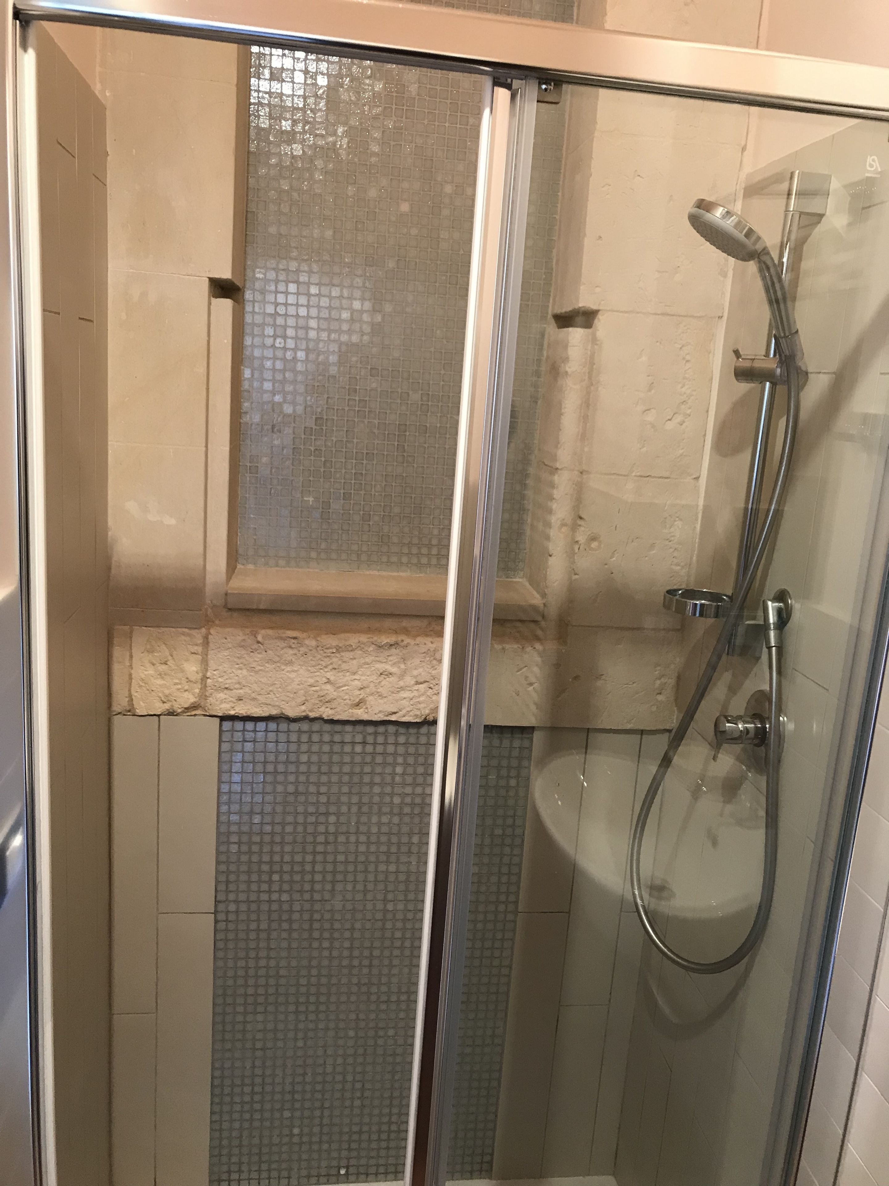 Um chuveiro com divisória de vidro, parede revestida com azulejos, janela e chuveiro manual.