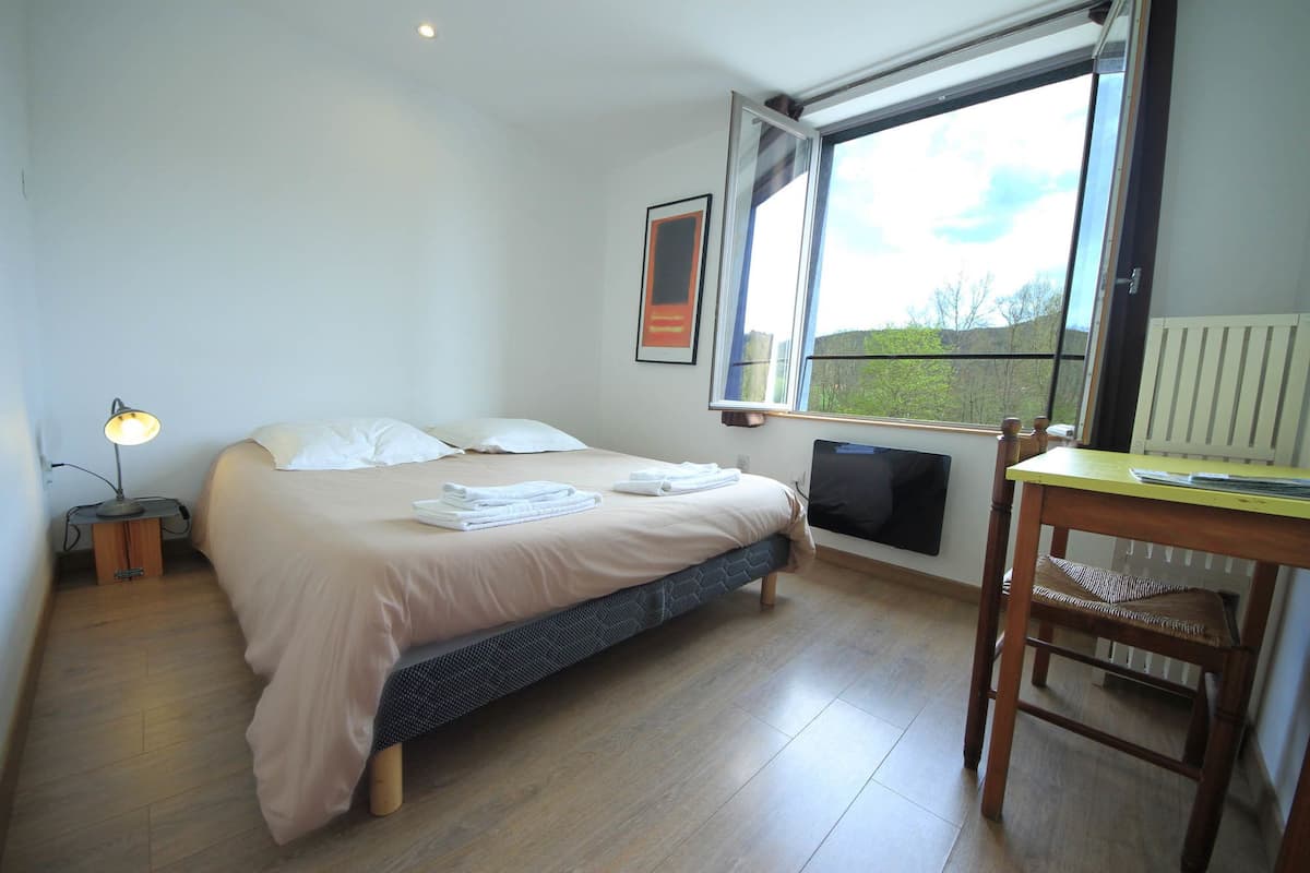 Quarto com cama, luminária de cabeceira, janela, radiador e uma pequena mesa.