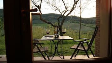 Uma mesa de jantar com quatro cadeiras, uma garrafa e dois copos de vinho, com colinas suaves e uma árvore ao fundo.