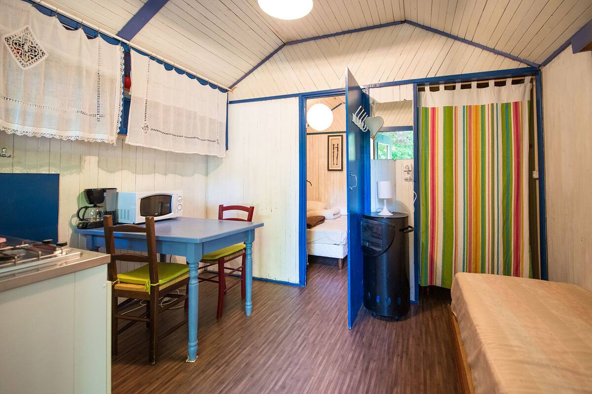 Quarto compacto com uma mesa de madeira, cadeiras, uma cama, uma lixeira e uma cortina listrada e colorida.