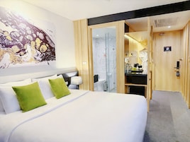Quarto de hotel com uma cama grande, box de vidro, guarda-roupa de madeira e uma pequena cozinha equipada.