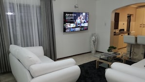 Uma sala de estar moderna com um sofá branco, uma mesa de centro e uma televisão exibindo um programa. É visível uma área de cozinha através de um arco.