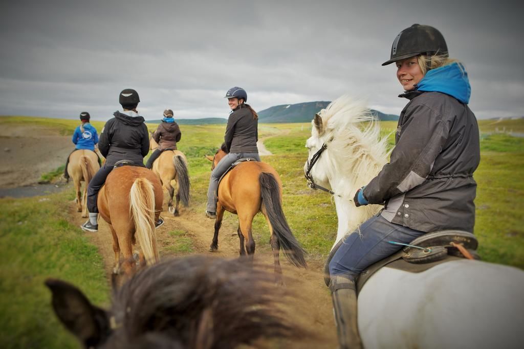 Um grupo de pessoas cavalgando em um caminho de terra em uma área gramada, com montanhas ao fundo.