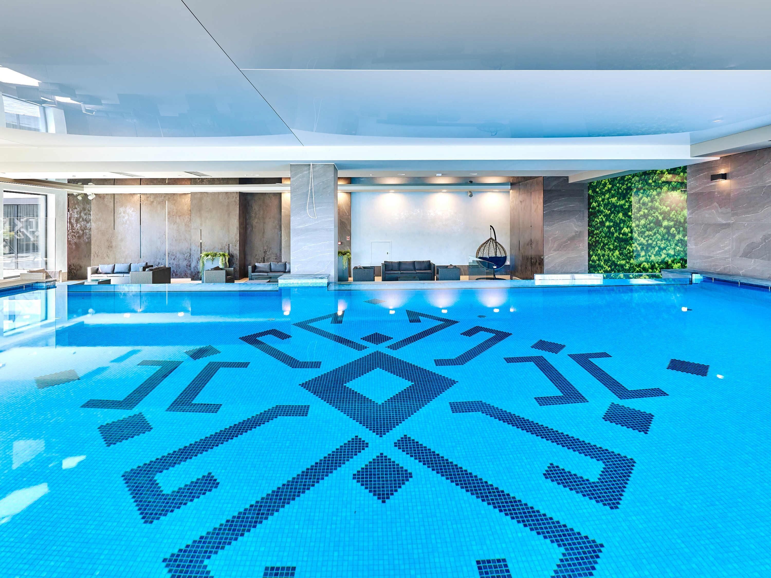 Piscina interior moderna com padrões geométricos de azulejos, área de estar com assentos e parede verde com folhagem.
