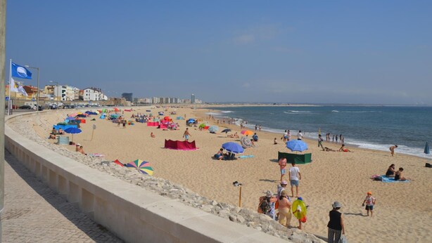 Uma praia com pessoas, guarda-sóis e edifícios ao fundo.