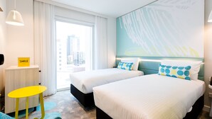 Quarto de hotel com duas camas, uma mesinha amarela, uma mesinha de cabeceira branca e uma janela grande com cortinas.