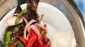 Um prato com um peixe inteiro, cebolas fatiadas, pimentões vermelhos e uma pequena tigela de molho.