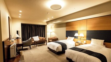 Quarto de hotel com duas camas, uma escrivaninha, uma cadeira, um abajur e uma janela grande com cortinas.