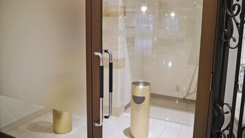 Entrada de um quarto de hotel com porta de vidro, puxador de metal, lixeira cilíndrica e um pequeno banquinho.