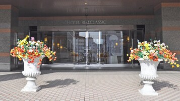 Entrada de hotel com porta de vidro, dois arranjos florais grandes e piso de azulejos.