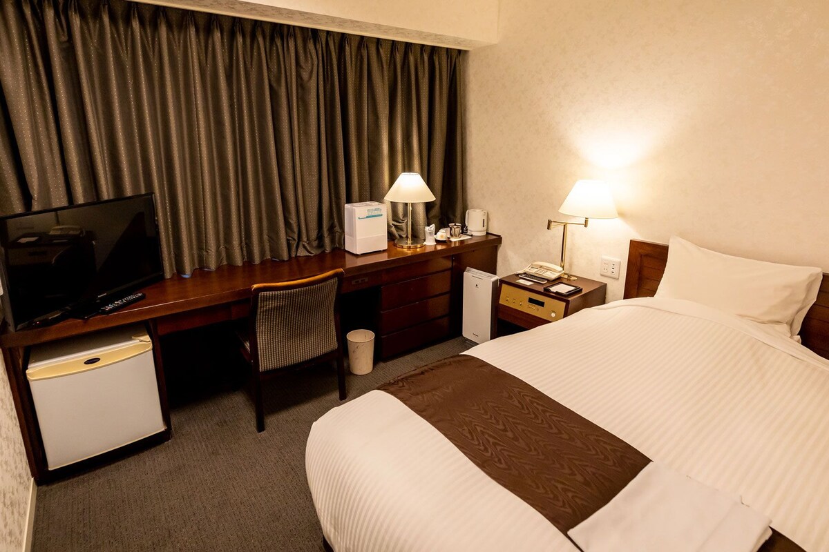 Quarto de hotel com cama, escrivaninha com televisão, cadeira, mesa de cabeceira com abajur e um pequeno frigorífico.