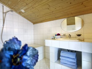 Casa de banho com teto de madeira, azulejos brancos, lavatório com espelho e chuveiro.