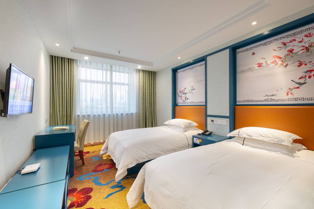 Quarto de hotel com duas camas, uma escrivaninha, uma cadeira, uma televisão e uma janela com cortinas.