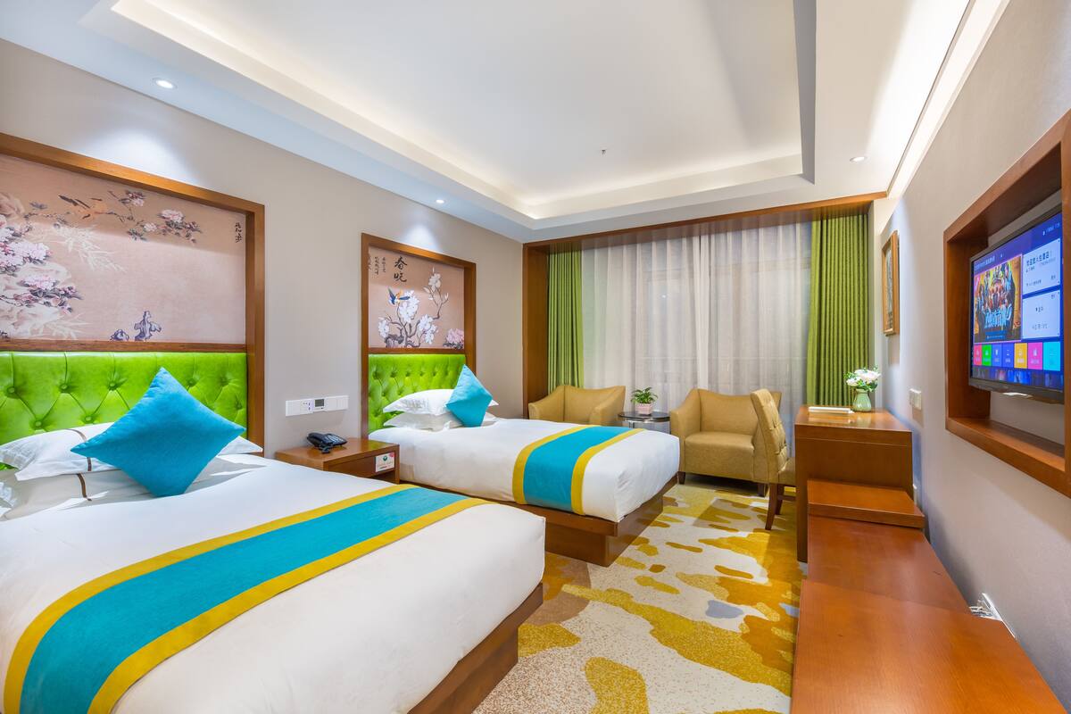 Quarto de hotel com duas camas, cabeceira estofada verde, televisão e uma grande janela com cortinas translúcidas.