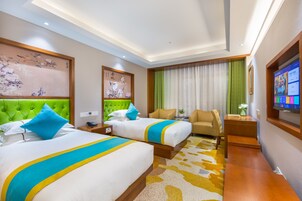 Quarto de hotel com duas camas, cabeceira estofada verde, televisão e uma grande janela com cortinas translúcidas.