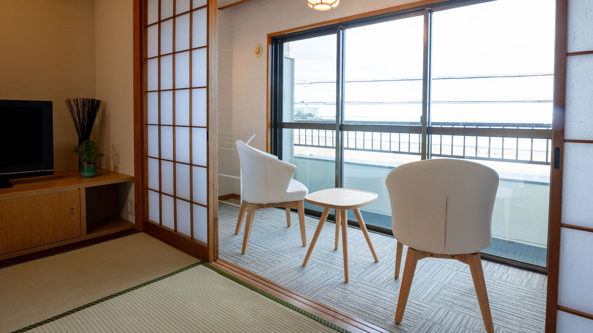 Quarto japonês tradicional com tatames, um painel deslizante shoji, uma televisão e duas cadeiras brancas perto de uma janela grande.