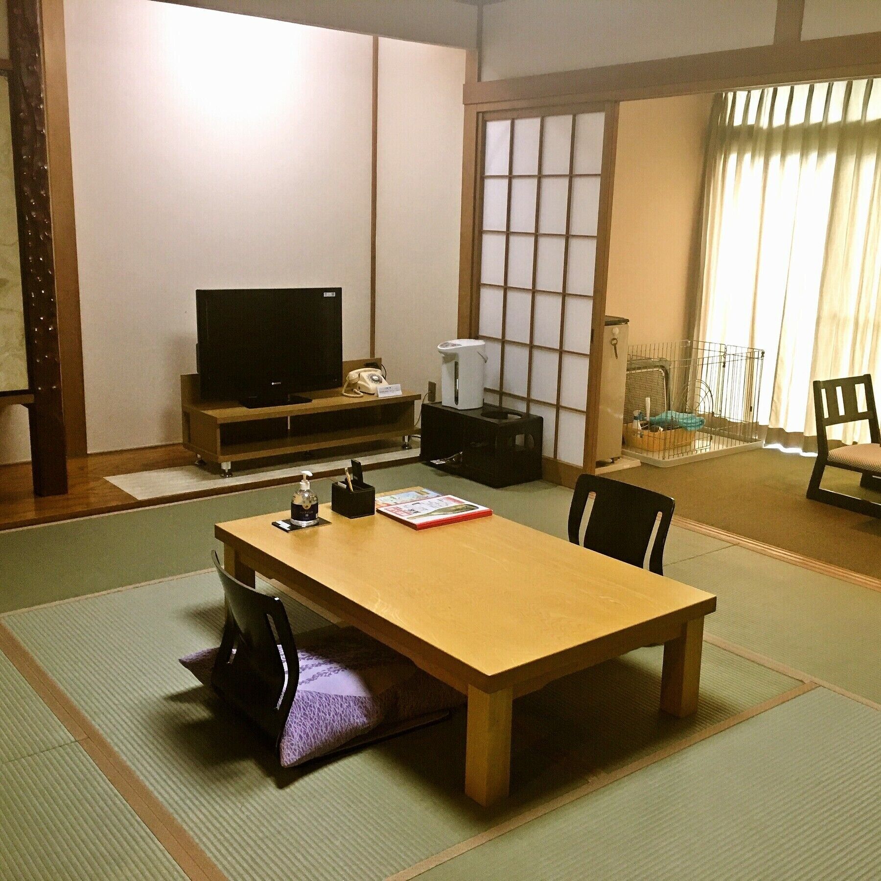 Um quarto tradicional em estilo japonês, com piso de tatame, uma mesa de madeira baixa, uma televisão e uma porta deslizante que dá para uma varanda.