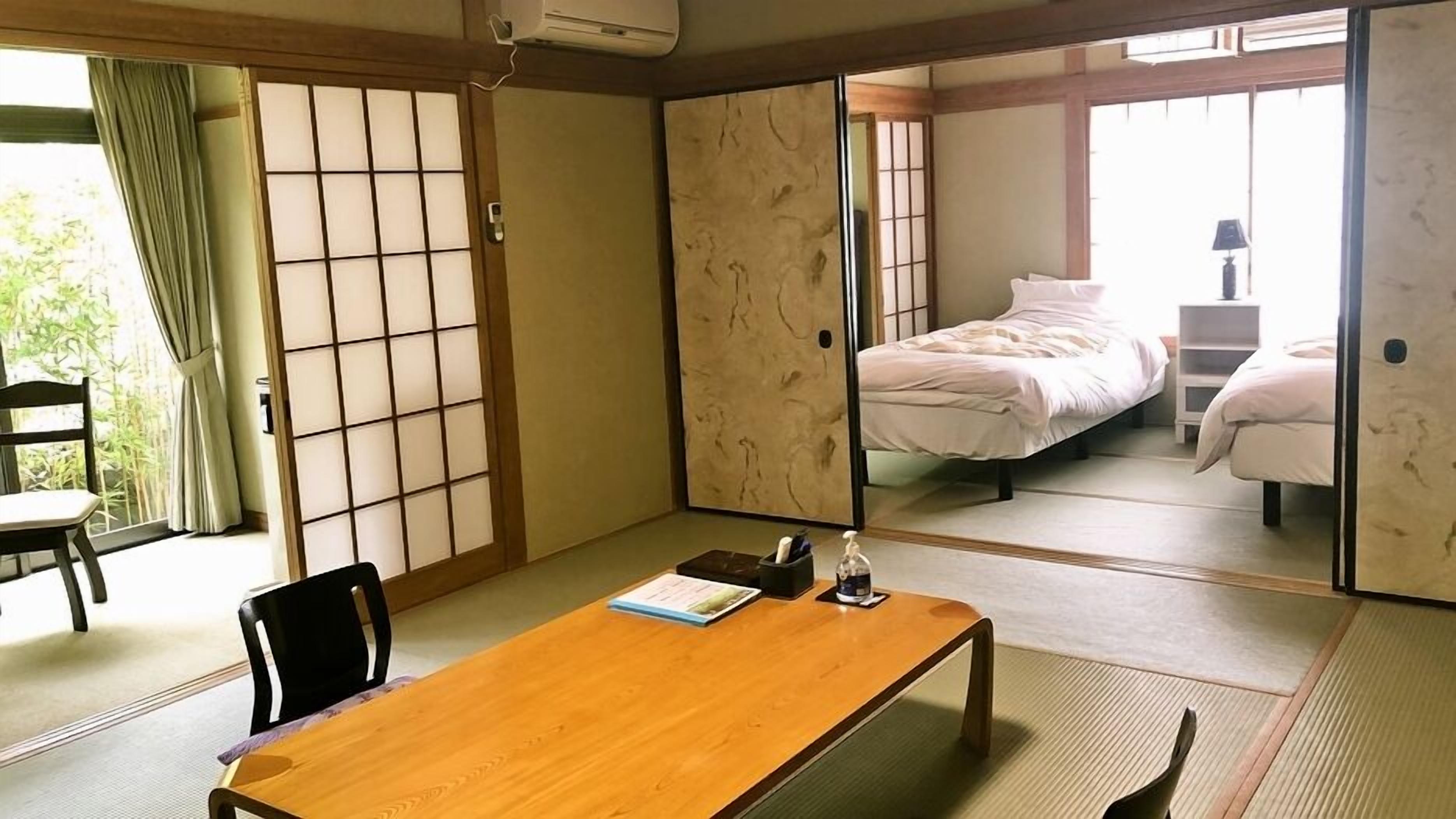 Um quarto japonês tradicional com tatames, uma mesa de madeira, uma cadeira e uma cama com colcha branca.