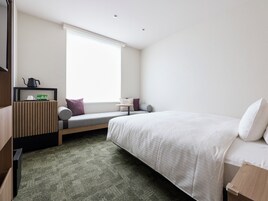 Quarto de hotel com uma cama, uma mesinha, um banco e uma janela.