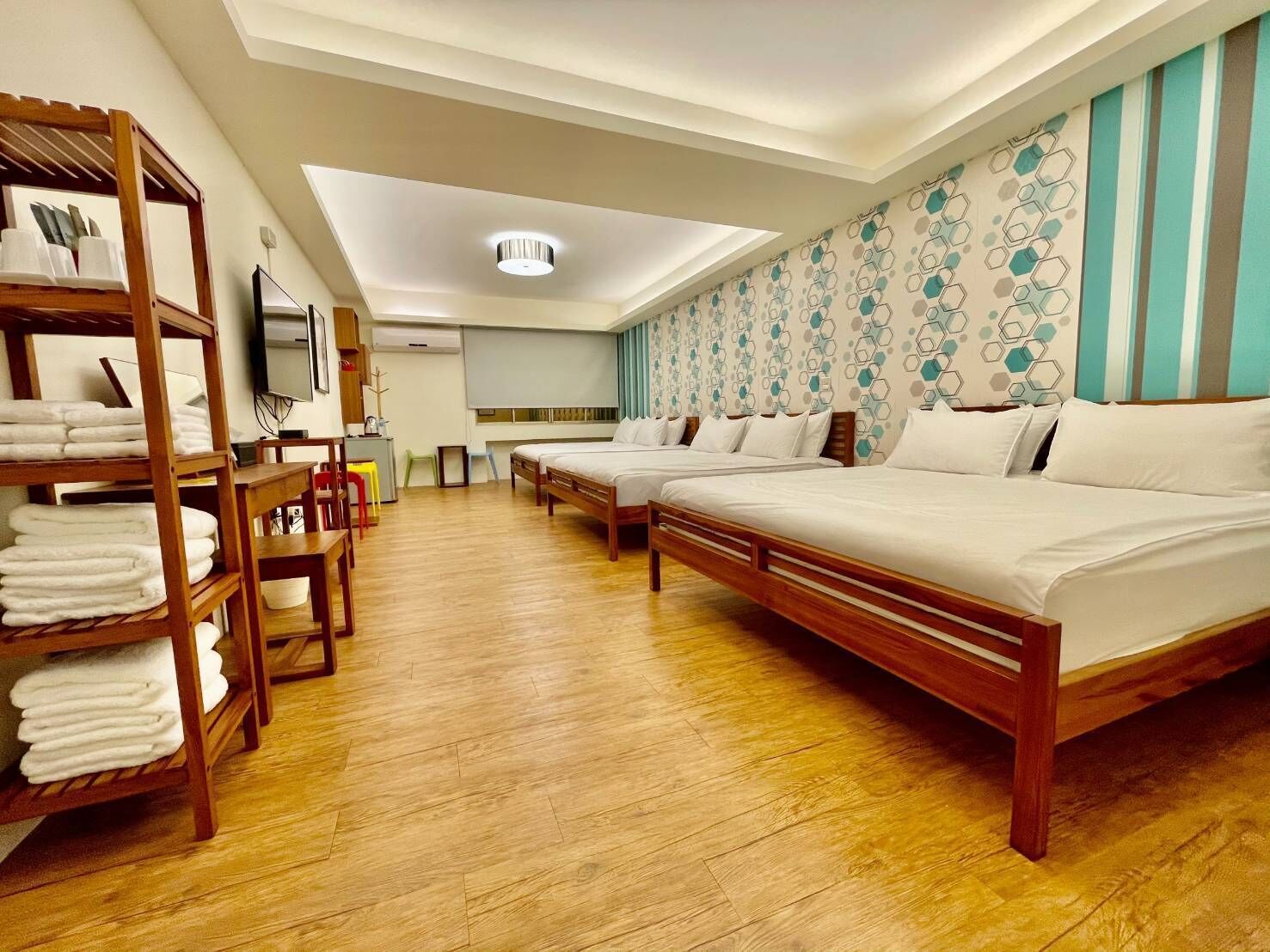 Quarto de hotel com uma cama grande, prateleira de madeira com toalhas, escrivaninha com cadeira e parede estampada.