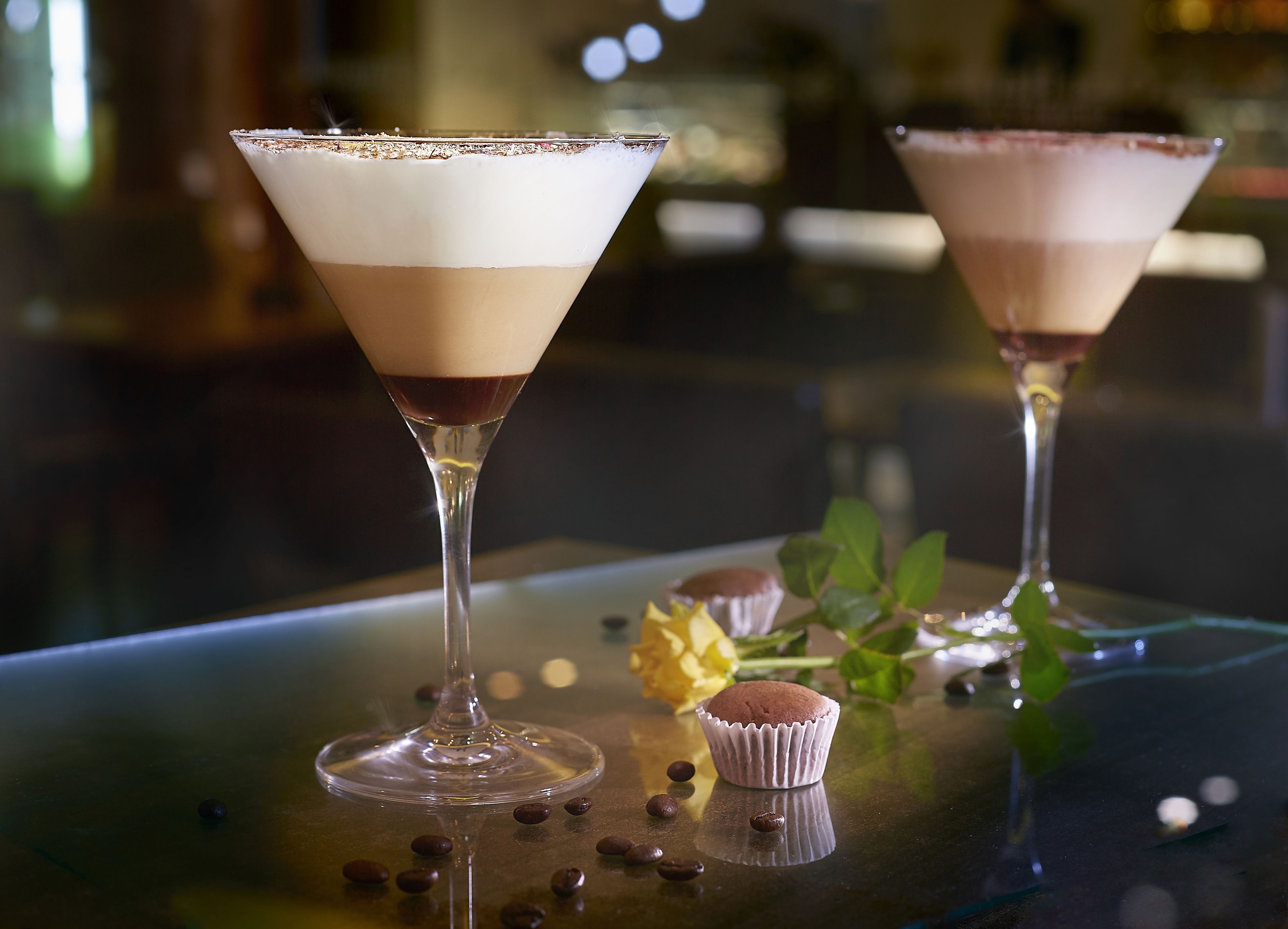 Dois cocktails em camadas em copos de martini sobre uma superfície refletora, com um cupcake e grãos de café.