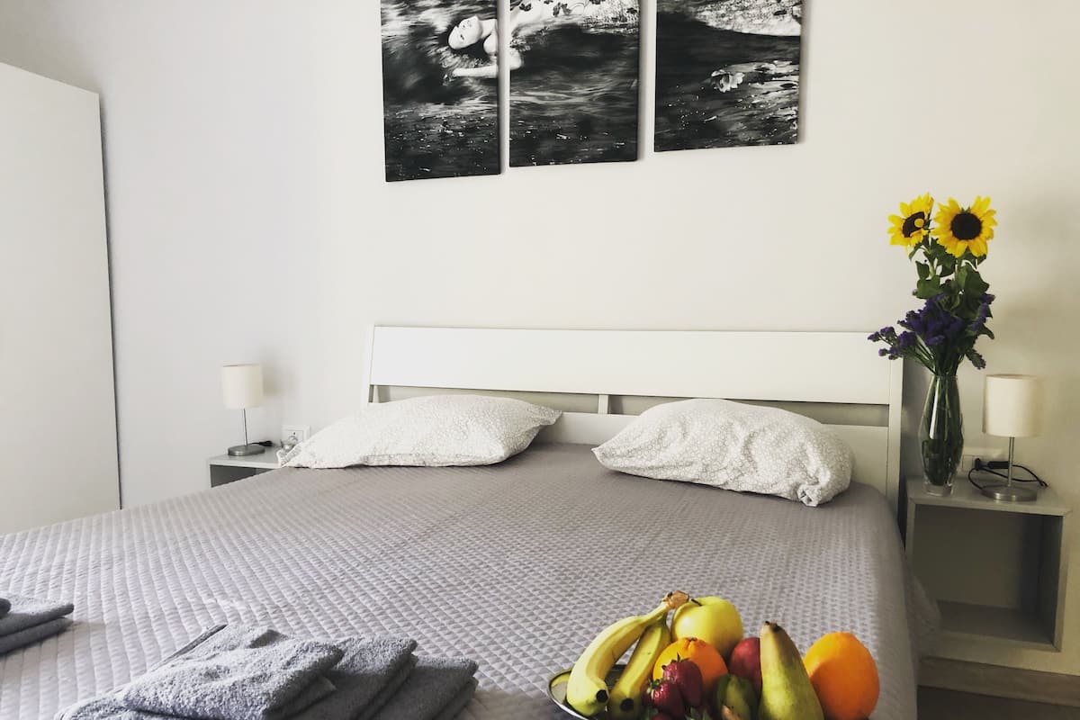 Cama bem arrumada com um edredom cinza, duas almofadas, uma mesinha de cabeceira com uma luminária, um vaso com flores e uma fruteira na cama.