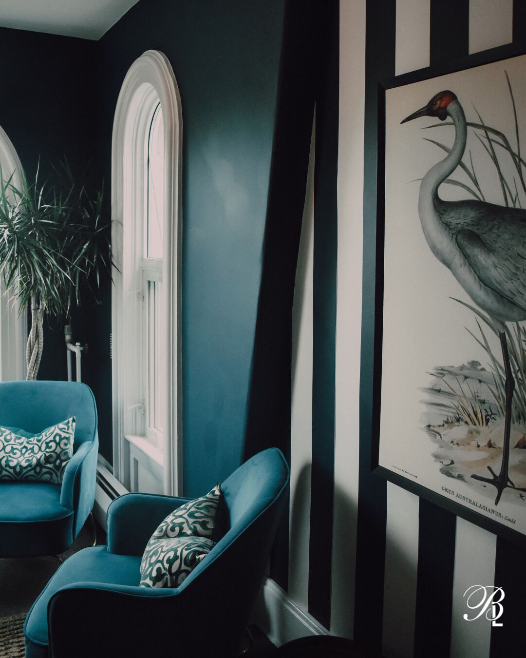 Quarto com parede azul, quadro de pássaros emoldurado, poltrona azul e janela com moldura branca.