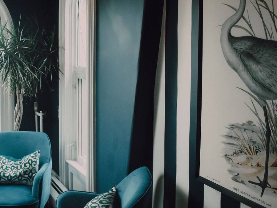 Quarto com parede azul, quadro de pássaros emoldurado, poltrona azul e janela com moldura branca.