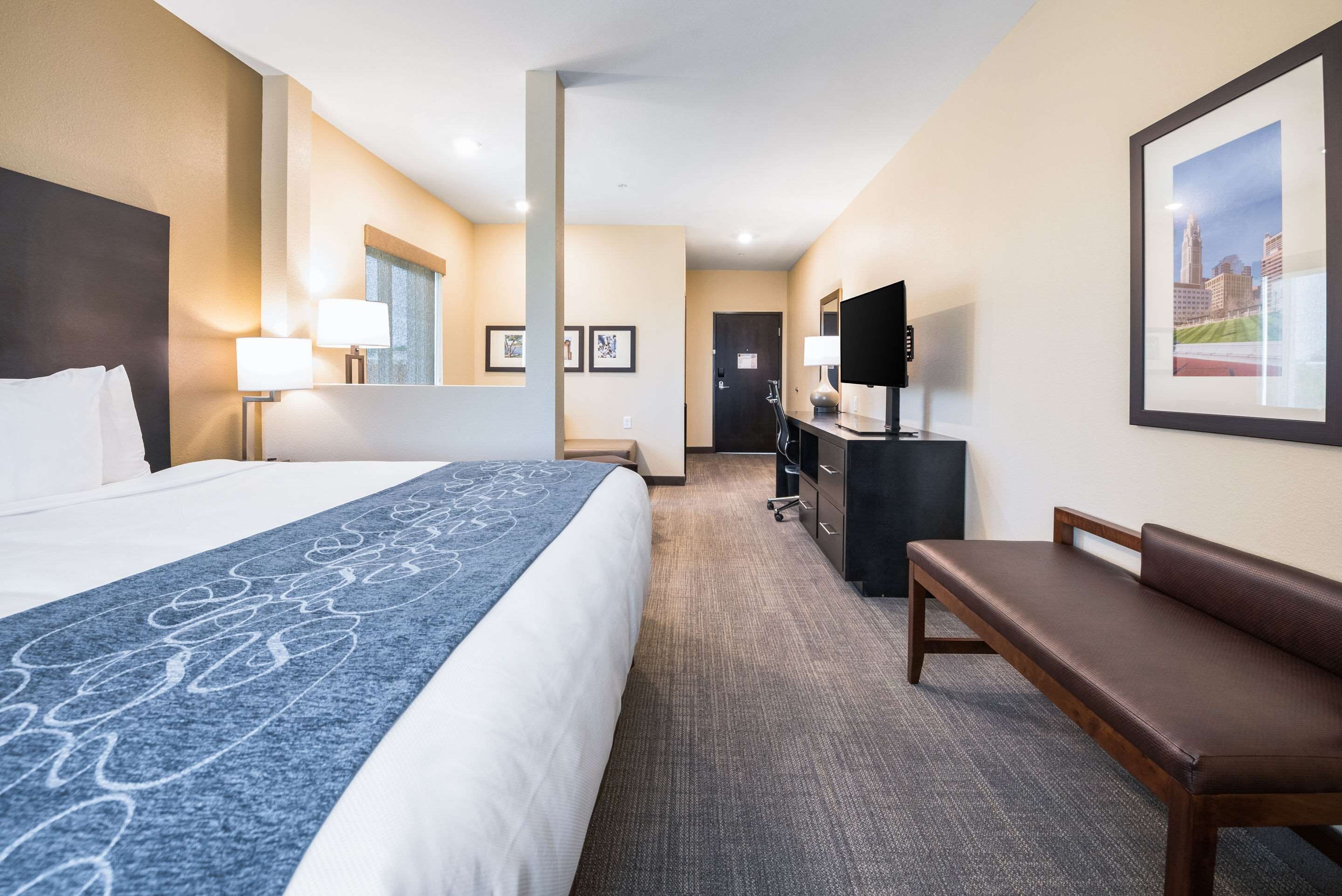 Quarto de hotel com uma cama grande, um banquinho, uma escrivaninha com televisão e quadros nas paredes.