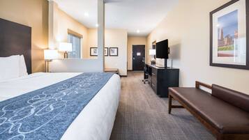 Quarto de hotel com uma cama grande, um banquinho, uma escrivaninha com televisão e quadros nas paredes.