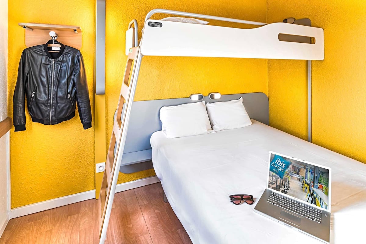 Quarto compacto com cama beliche, uma jaqueta pendurada, um laptop e um par de óculos de sol sobre a cama.