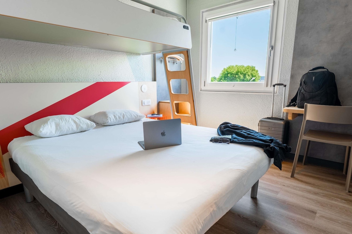 Quarto de hotel moderno com uma cama, um laptop, uma mala e vista para áreas verdes lá fora.