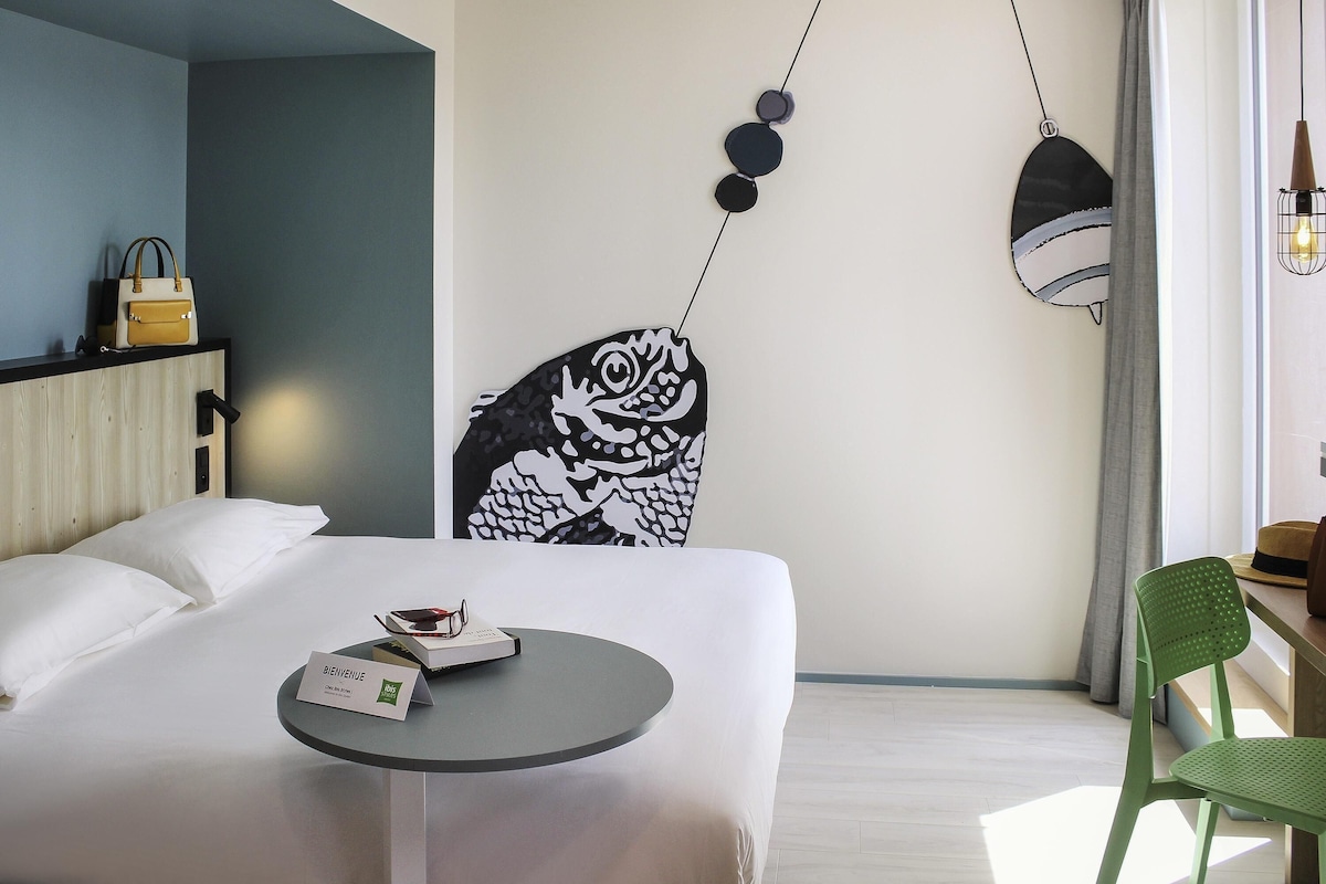 Quarto de hotel com uma cama, uma pequena mesa redonda, uma cadeira verde, uma obra de arte na parede e um lustre.