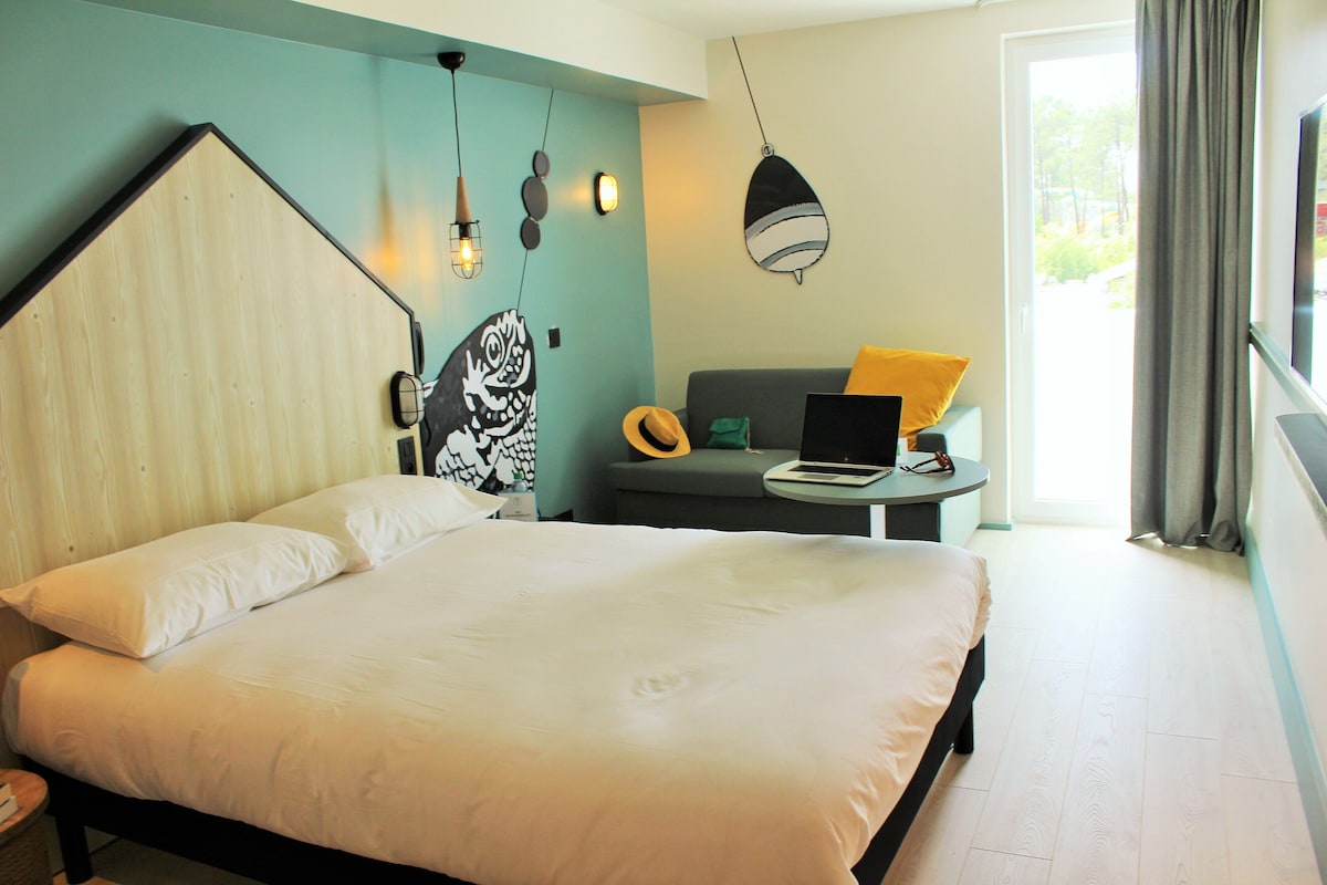 Quarto moderno com uma cama grande, uma escrivaninha com um laptop e vista para o exterior.