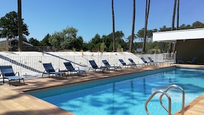 Uma piscina com deck de madeira e espreguiçadeiras, cercada por árvores altas e um muro branco.