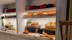 Vitrine de padaria com diversos pães e doces, um quadro negro com o cardápio e uma planta.