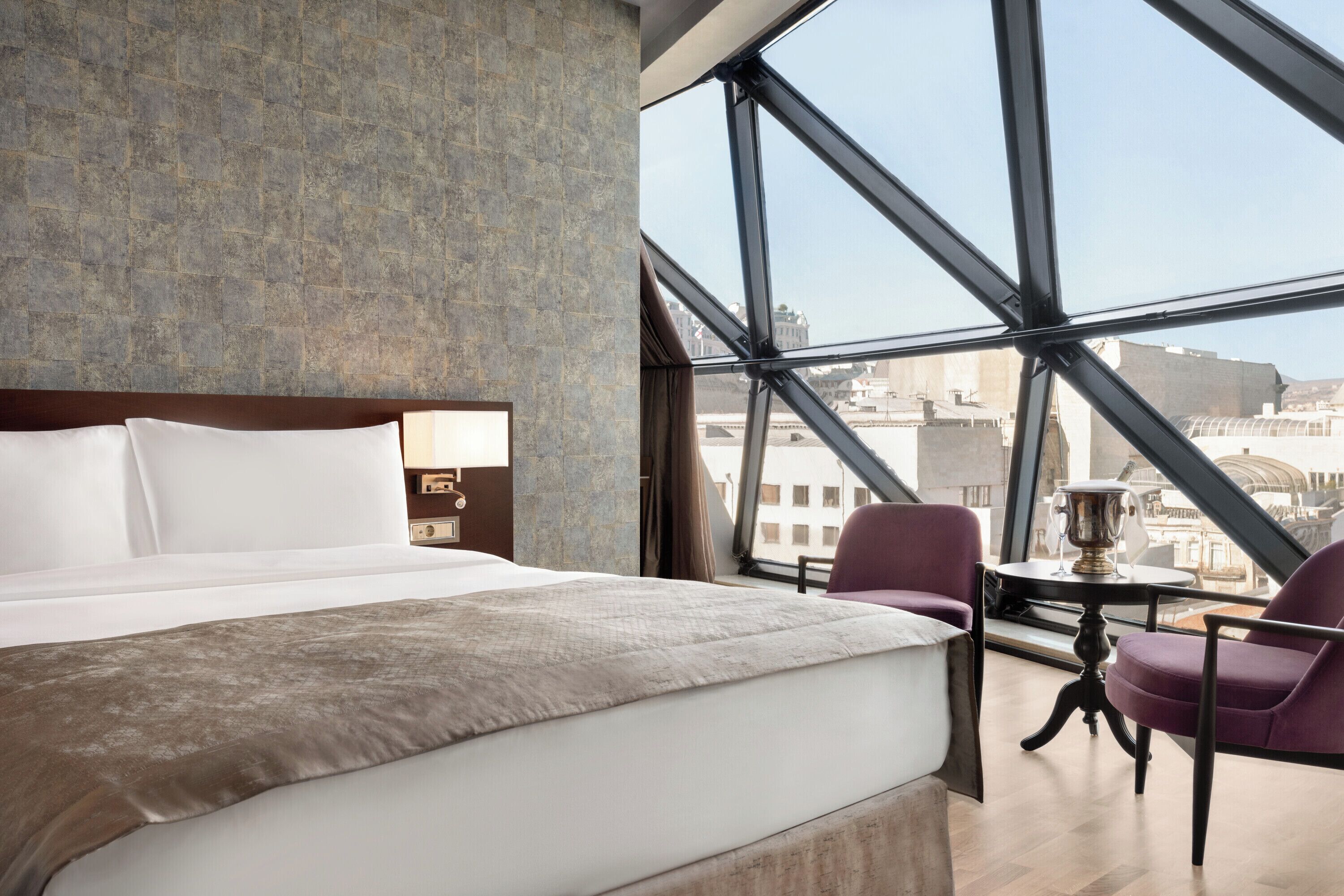 Quarto de hotel com uma cama grande, vista através de uma janela ampla e uma pequena mesa com luminária.