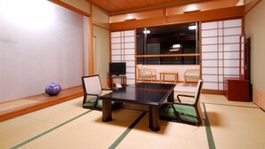 Um quarto japonês tradicional com tatames, uma mesa baixa com almofadas, uma televisão e biombos shoji.