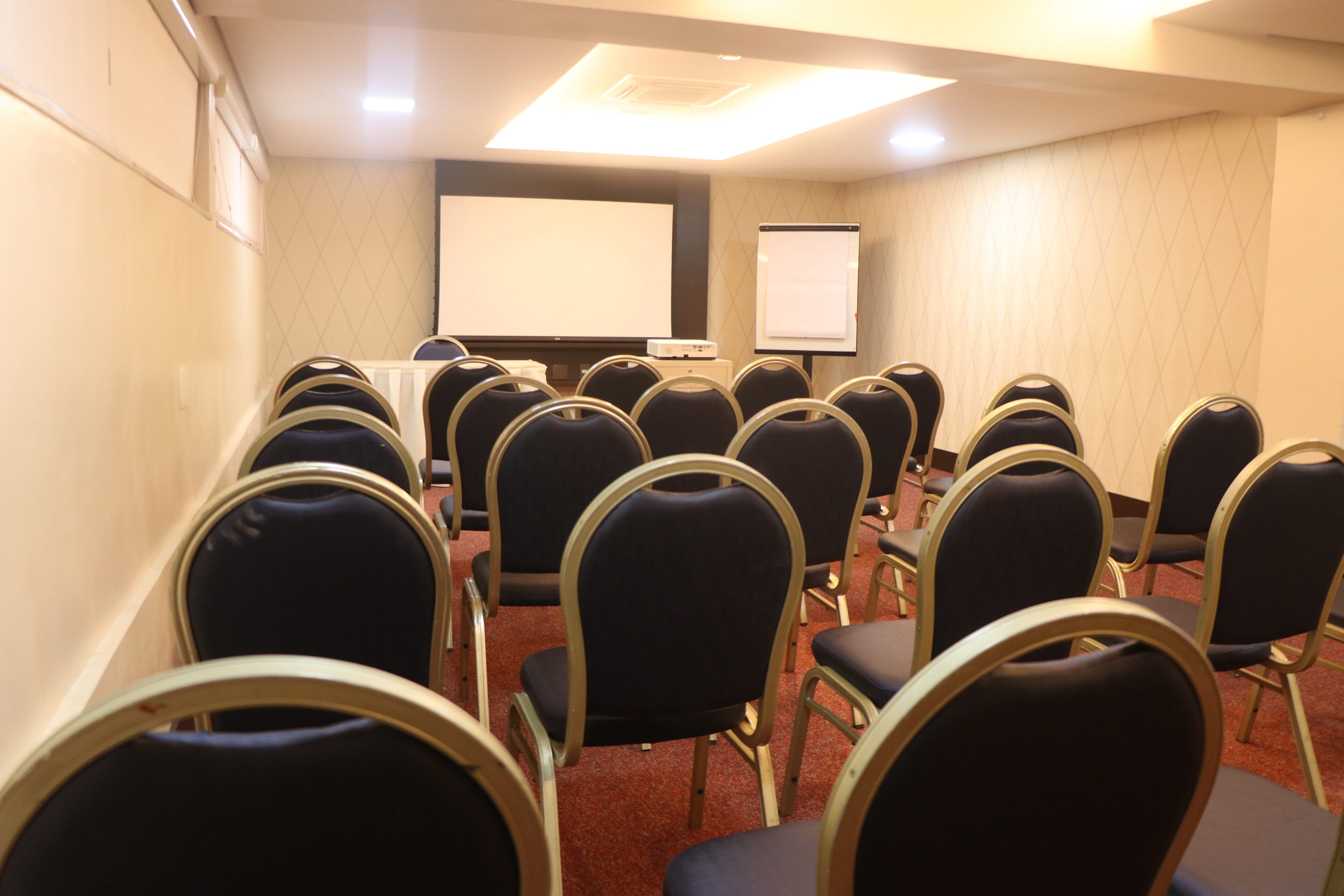 Uma sala de conferências com fileiras de cadeiras, uma tela de projetor e um quadro branco.