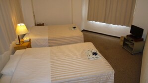 Quarto de hotel com duas camas, um relógio na parede, uma televisão sobre um suporte e uma janela com cortinas.