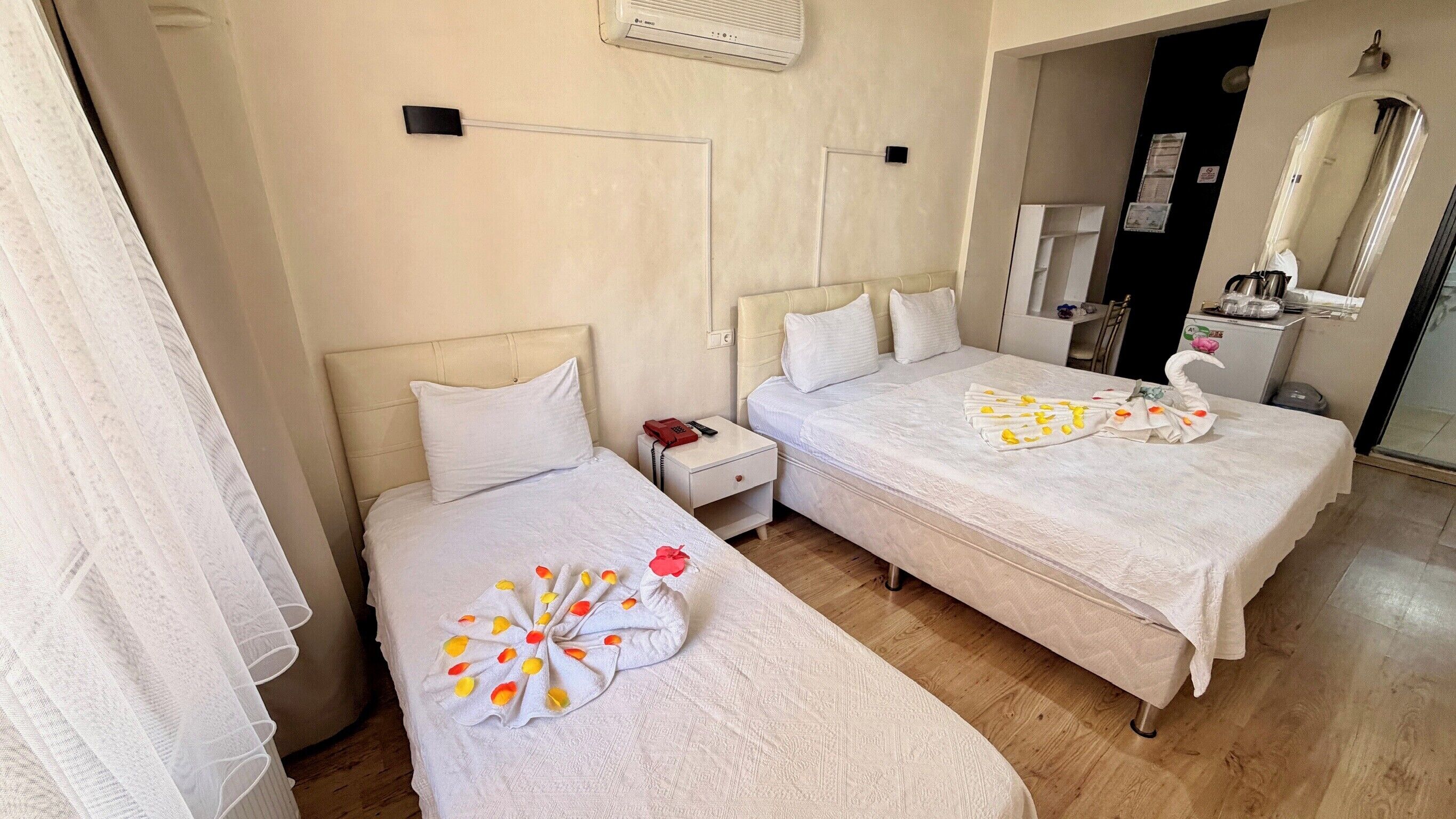 Quarto de hotel com duas camas, uma mesinha de cabeceira, um espelho, uma pequena cozinha e uma porta que dá para outro cômodo.