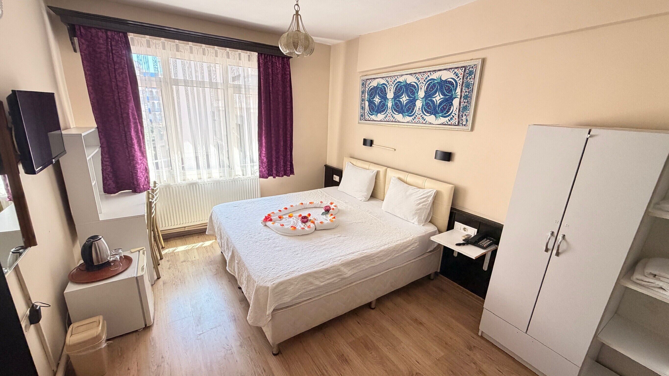 Quarto de hotel com cama, mesa de cabeceira com telefone, armário, janela com cortinas e uma obra de arte na parede.