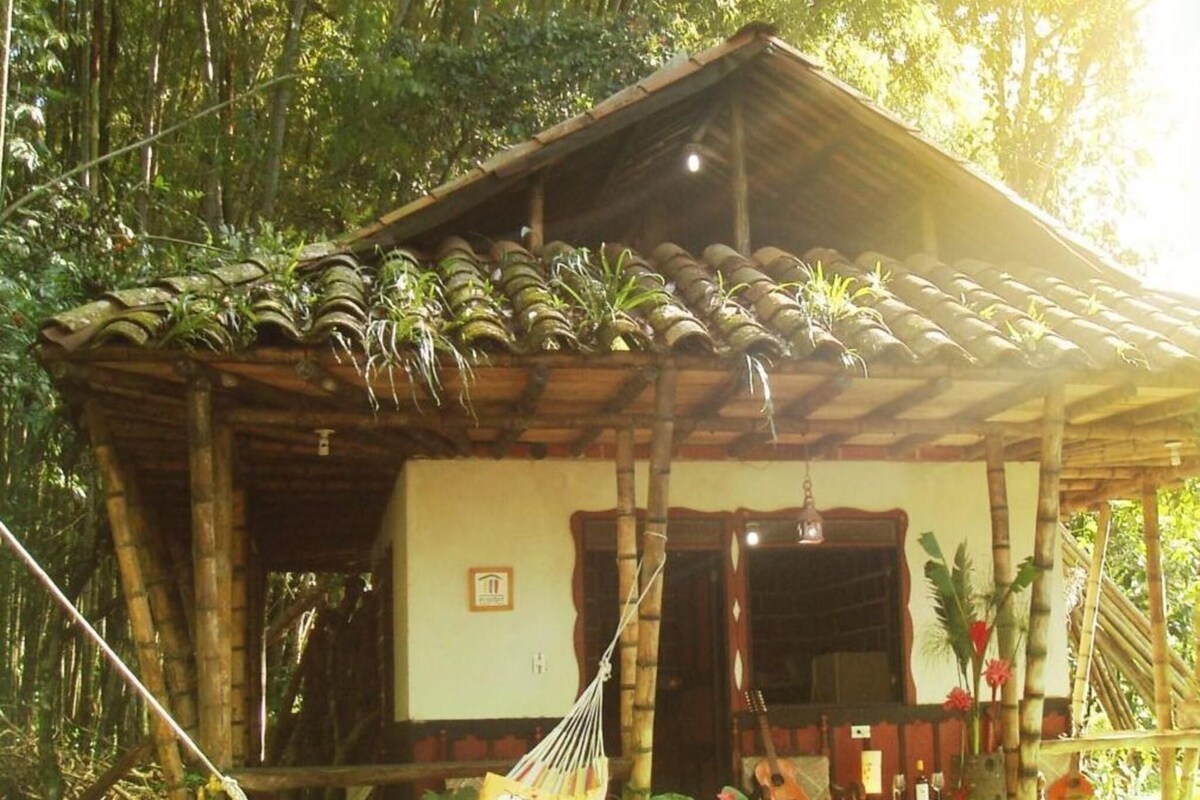 Uma casa rústica de madeira com telhado de palha, uma rede e um pequeno alpendre.