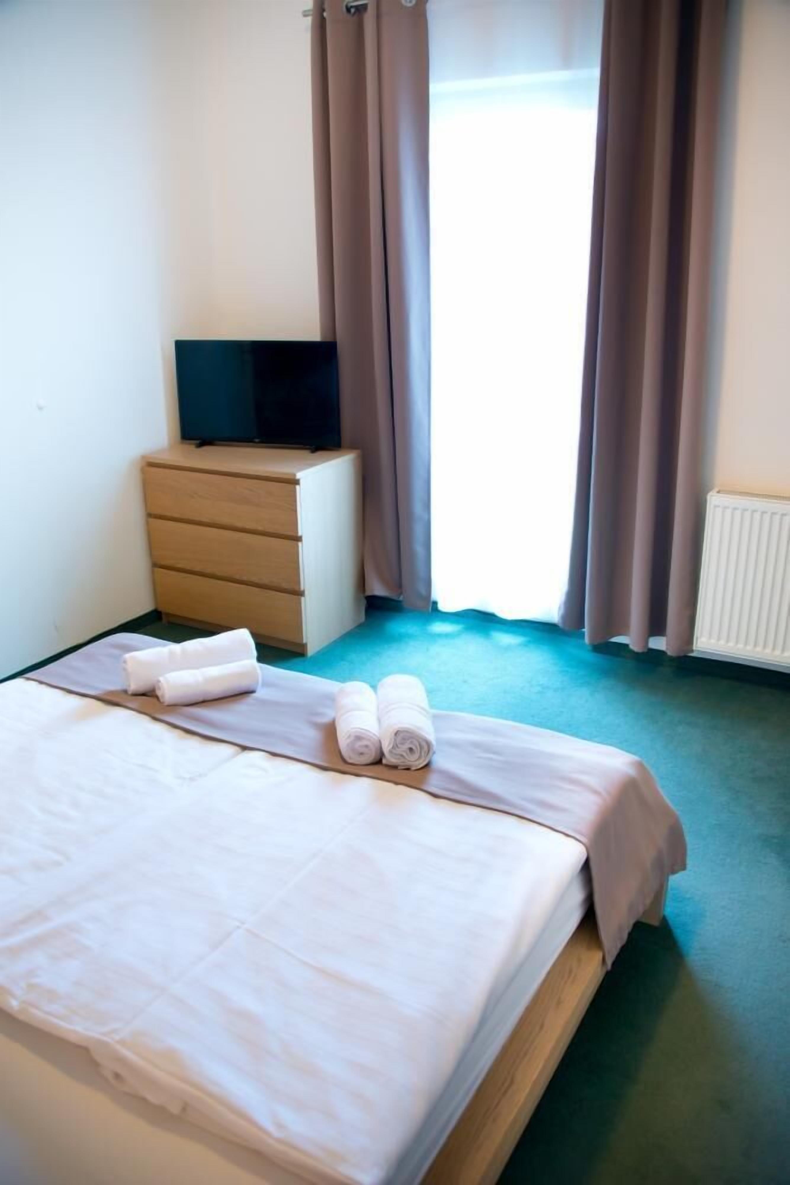 Quarto de hotel com uma cama, uma televisão sobre uma cômoda de madeira e uma janela com cortinas.