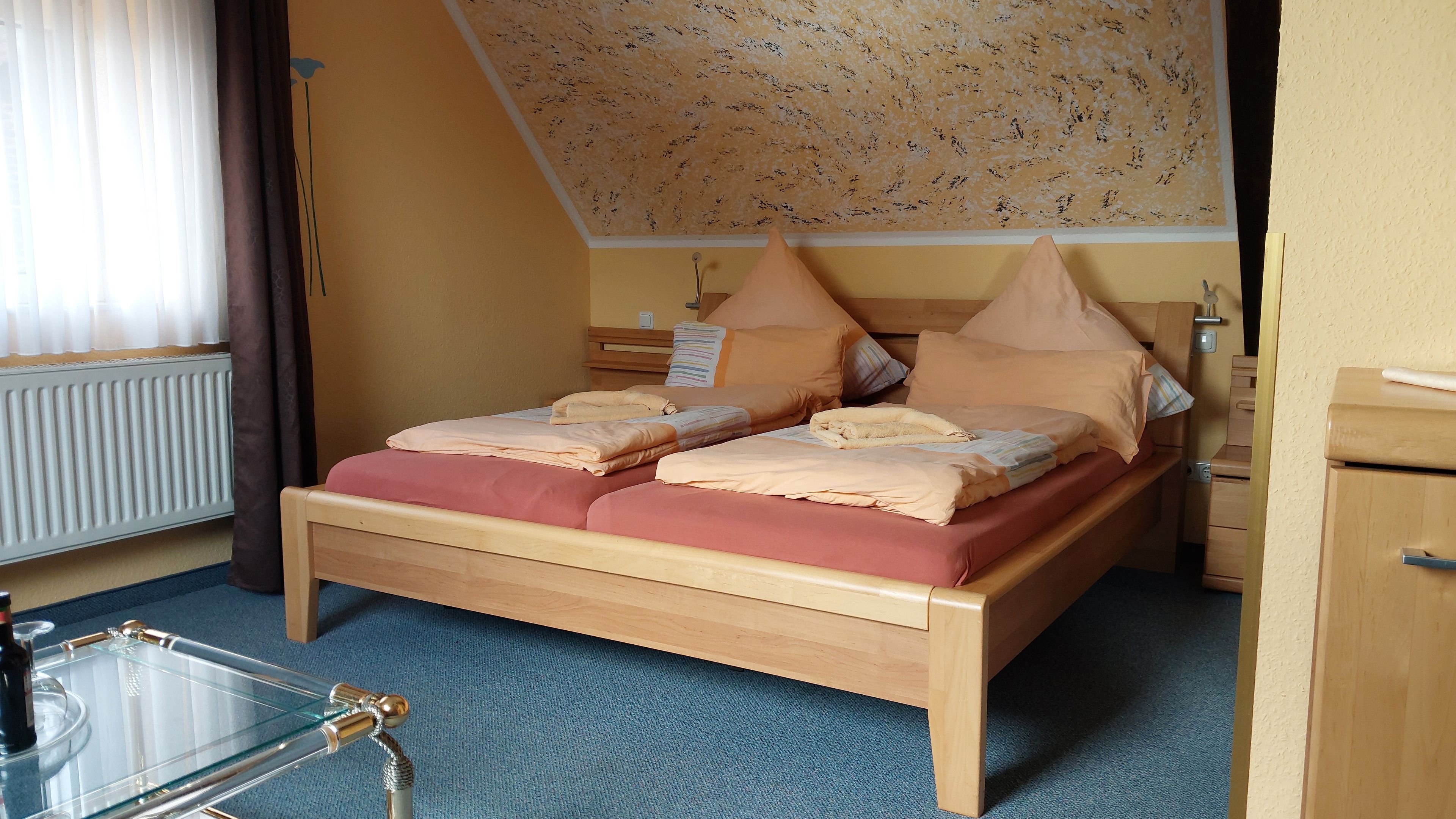 Uma cama de solteiro com estrutura de madeira, colchão vermelho e várias almofadas. Há uma mesa de cabeceira com abajur, uma janela com cortinas e um radiador.
